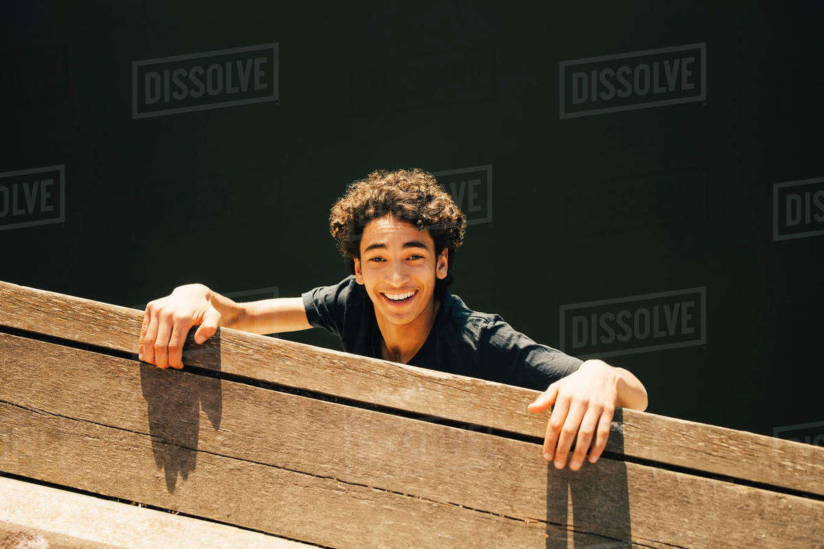 High angle portrait of cheerful teenage boy hanging from pier against ...