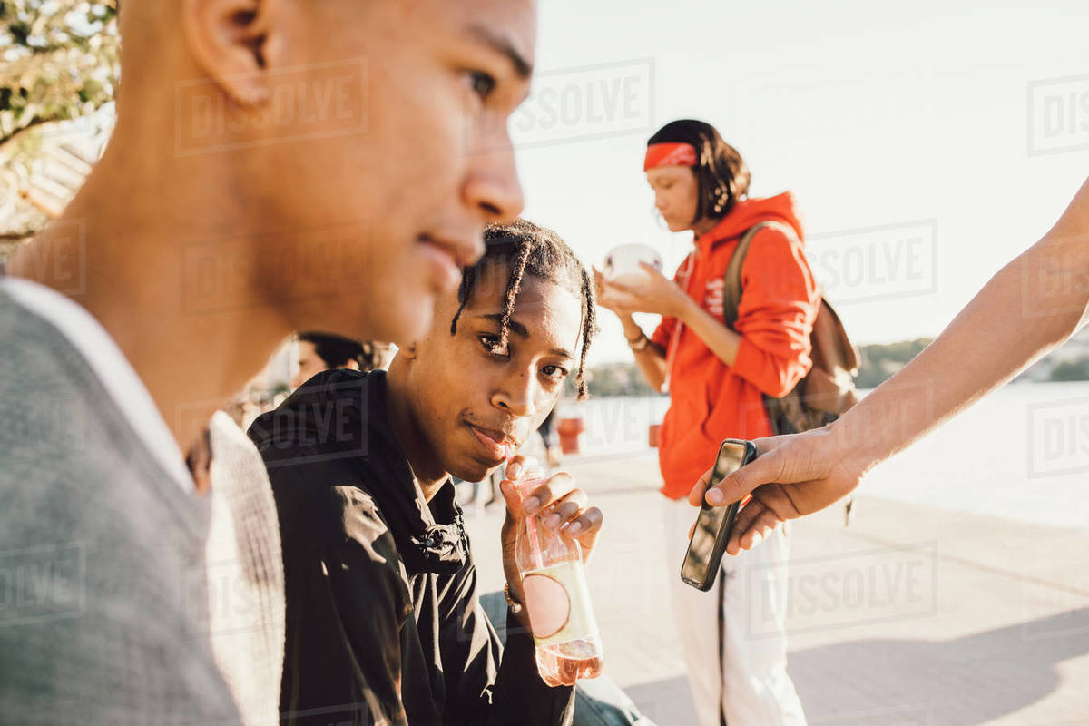 Portrait of teenage boy drinking drink while sitting with friends on ...