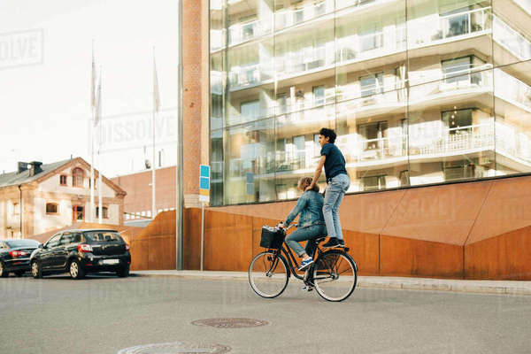 Side view of young man riding bicycle with friend on street in city ...