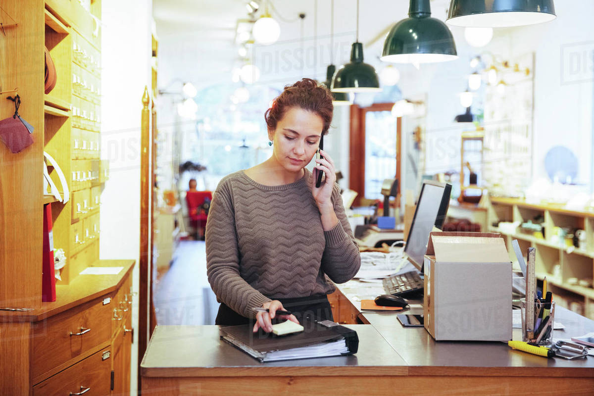 Young saleswoman talking on mobile phone standing at checkout in home ...