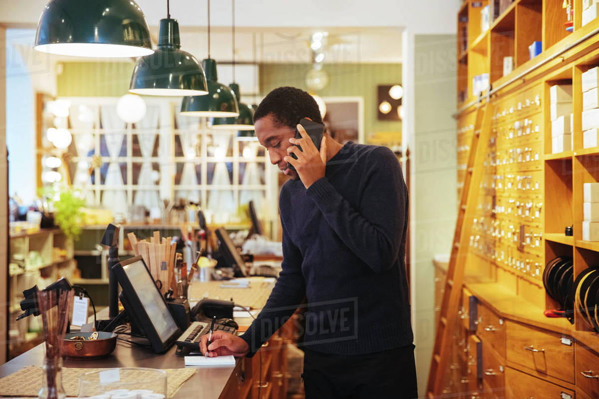 Confident young male owner talking on smart phone while writing at ...