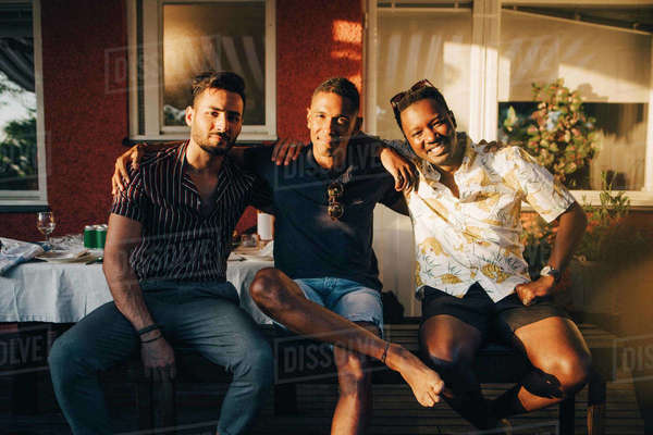Portrait of smiling male friends sitting with arms around at back yard ...