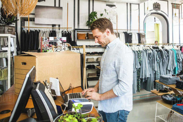 Side view of salesman using laptop while standing in clothing store ...