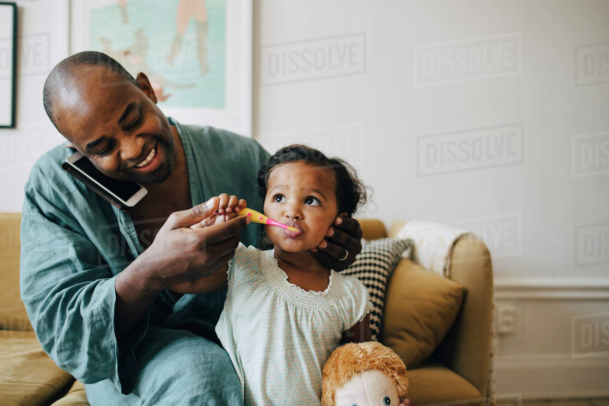 Man talking on mobile phone while brushing daughter's teeth at home ...
