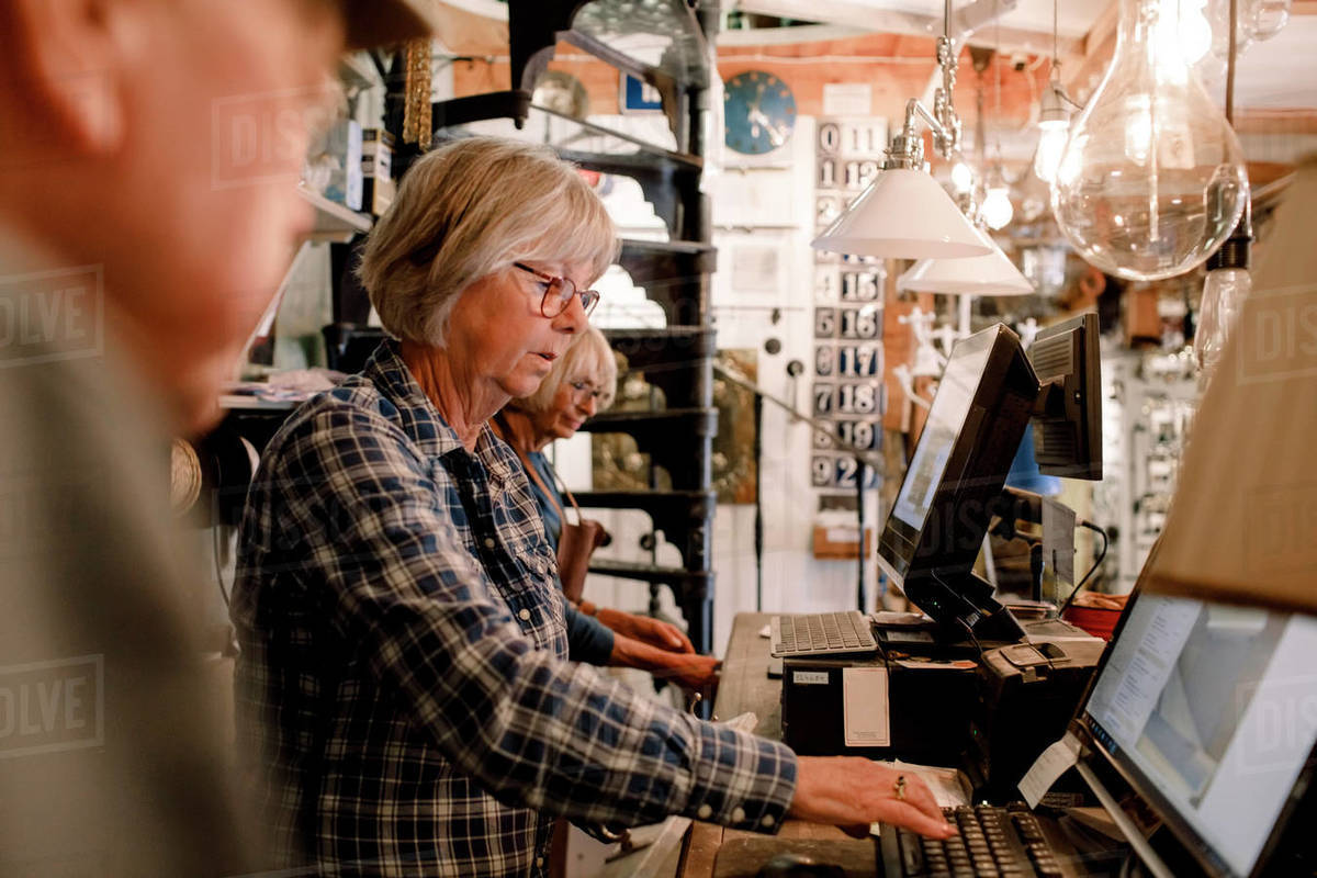 Confident senior female owner using computer while standing amidst ...