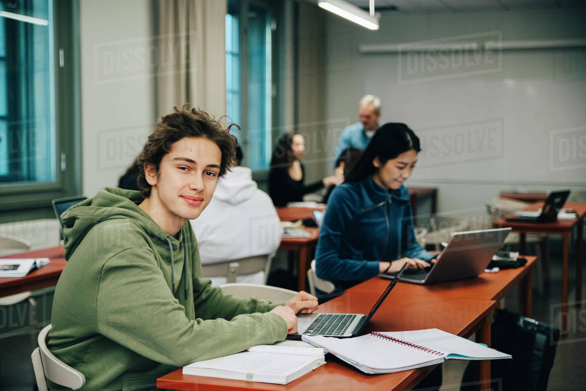 Portrait of smiling teenage boy using laptop at desk with classmates ...