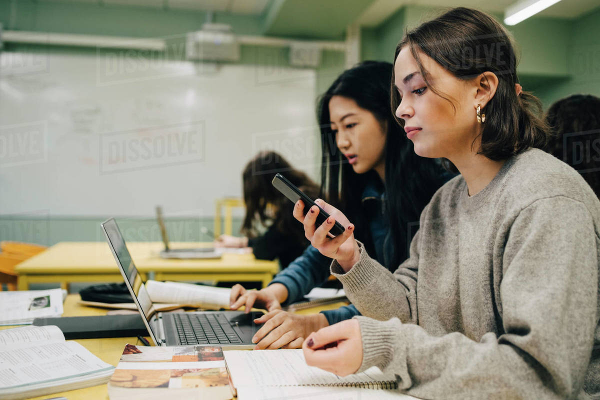 Female high school student using smart phone while sitting by classmate ...