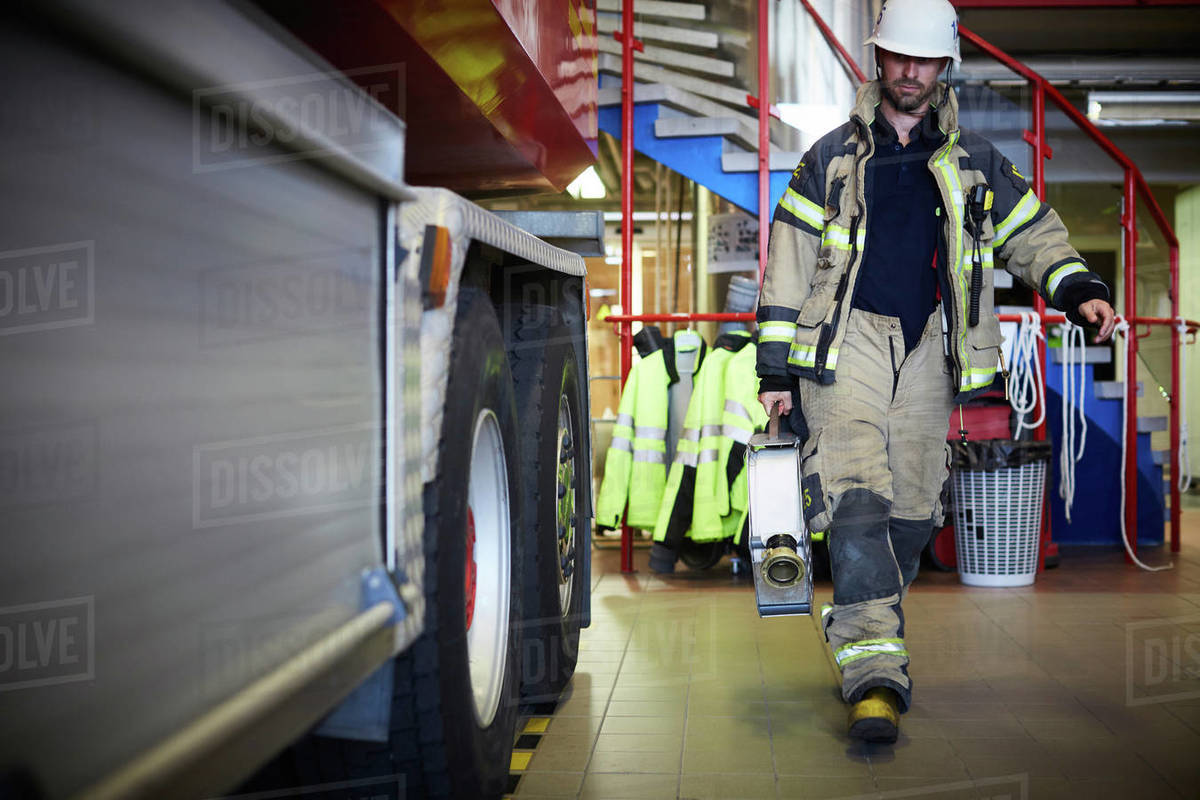 Full length of firefighter holding fire hose while walking by fire ...