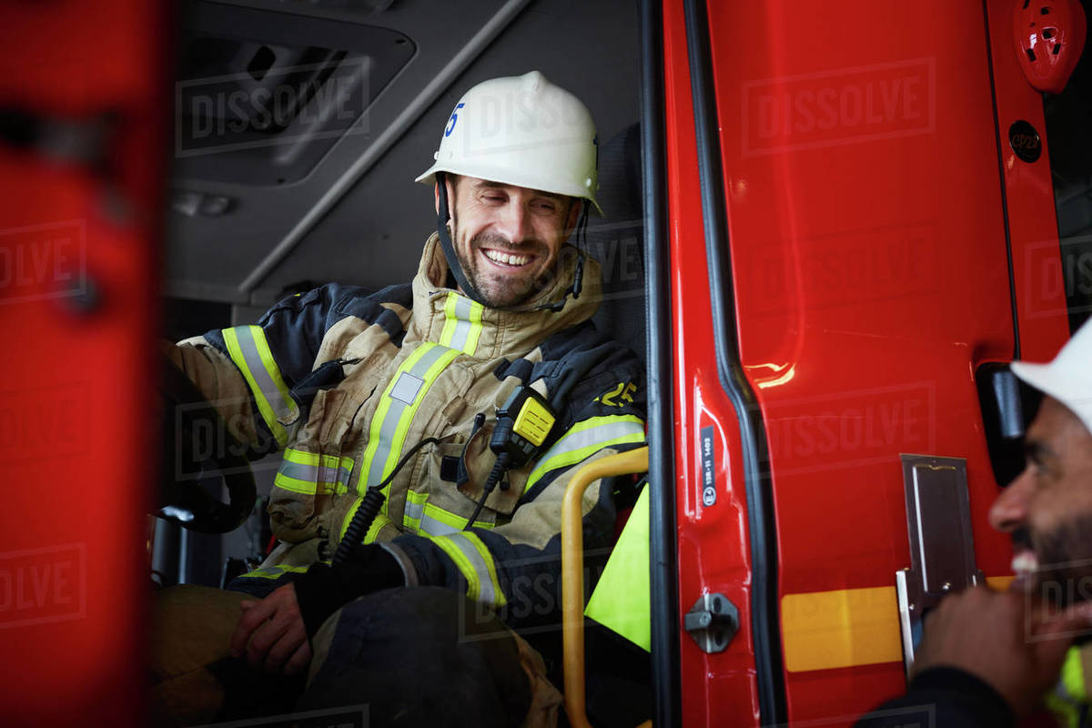 Smiling firefighter sitting in fire engine while talking to coworker ...