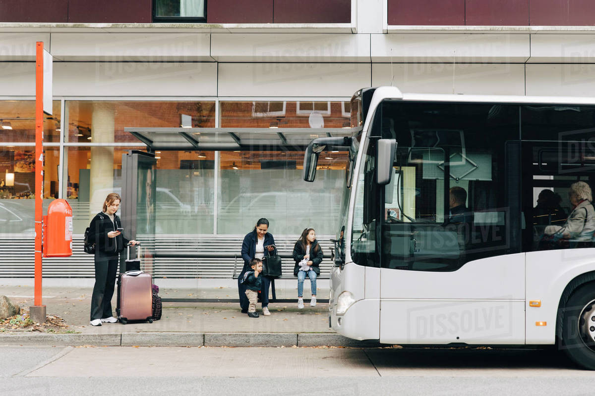 Full length of woman with luggage and family waiting for bus at bus ...