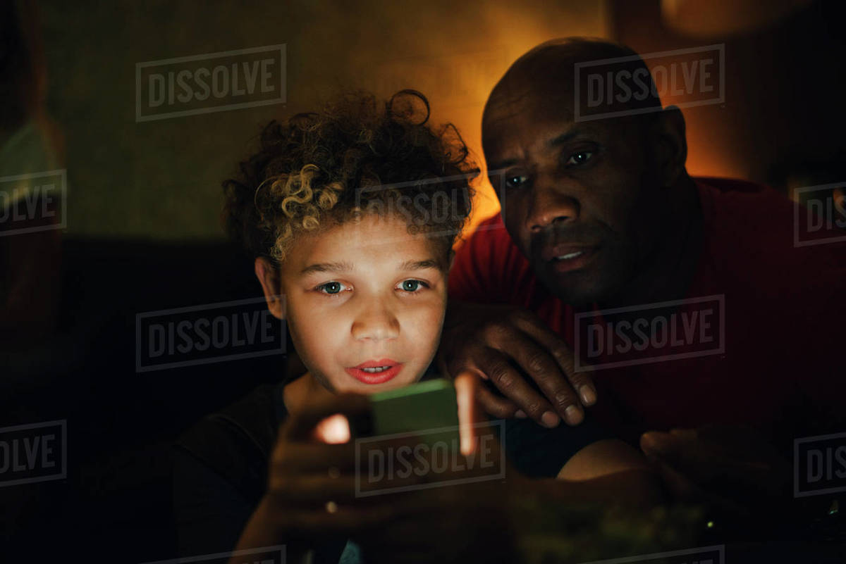 Boy using smart phone by father in living room at night - Royalty-free ...