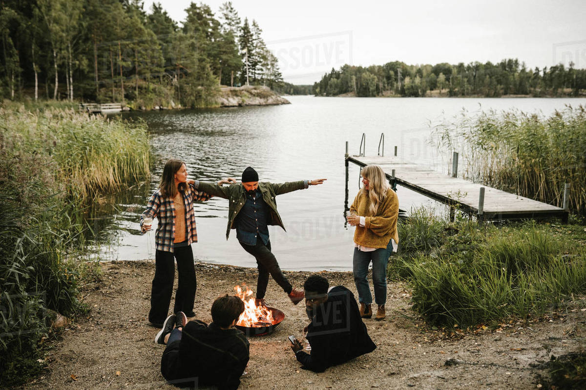 Male and female dancing by fire pit while friends sitting near lake ...