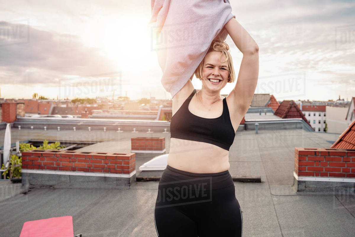 Portrait of smiling woman removing t-shirt on rooftop against dramatic ...