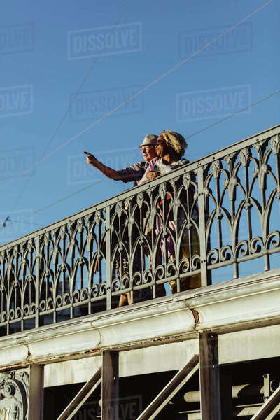 Senior man pointing while standing by woman on bridge against blue sky ...