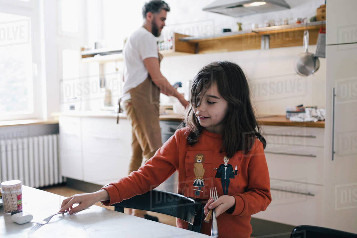 Girl arranging fork on dinging table while father doing chores in ...