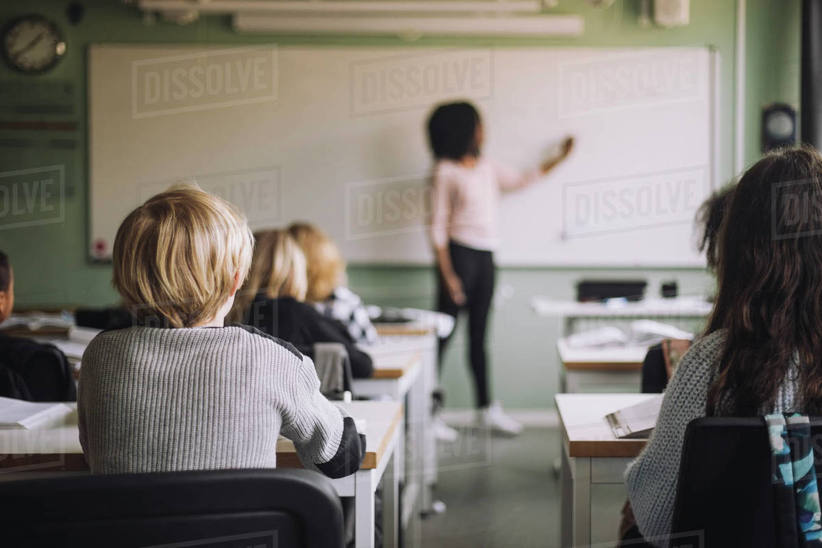 Rear view of students sitting at desk while teacher teaching in ...