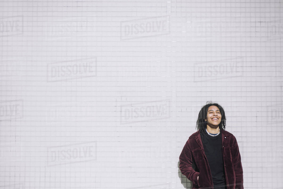 Cheerful genderqueer person standing with hand in pocket against wall ...
