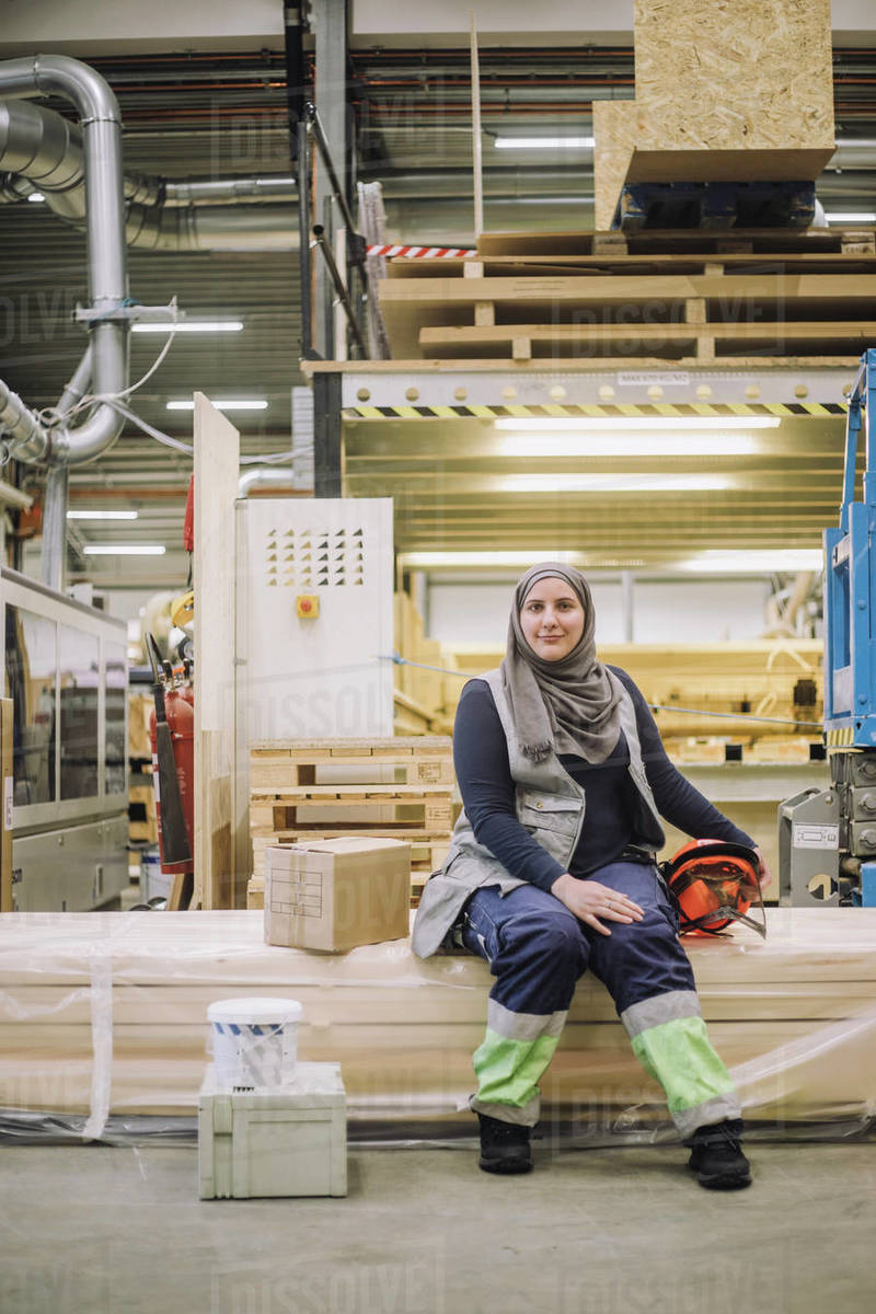 Portrait of female carpenter sitting with hardhat in warehouse ...