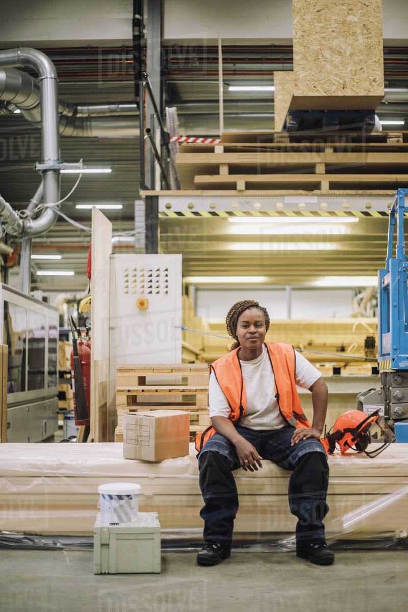 Portrait of female blue-collar worker sitting by hardhat in warehouse ...