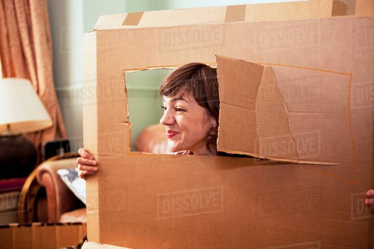 Mid adult woman peeking out of cardboard box window in living room ...