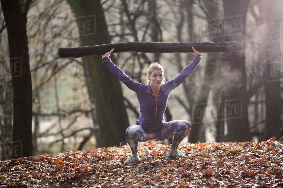 Front view of young woman squatting in forest arms raised holding tree ...