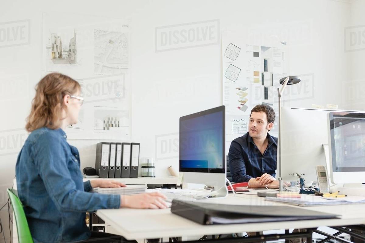 Colleagues sitting at desk in office using computers talking - Stock ...