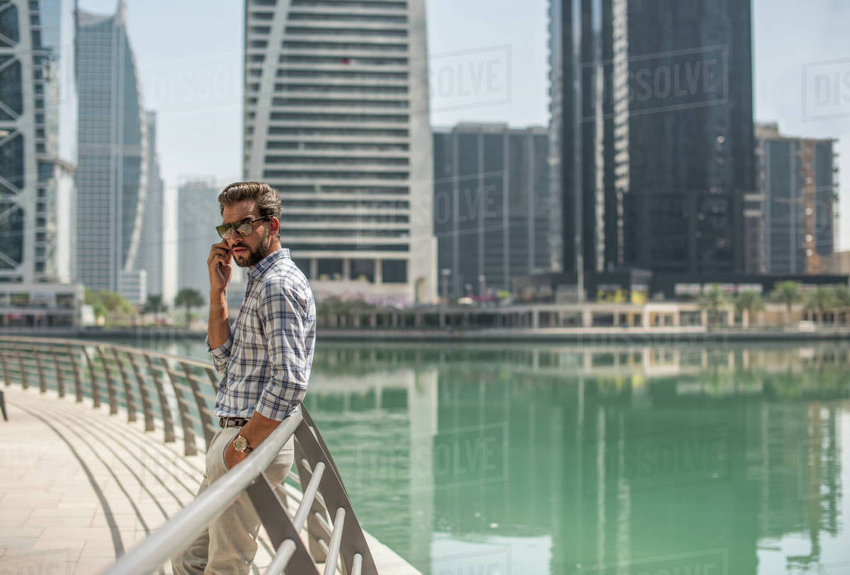 Young man leaning against waterfront railings talking on smartphone ...