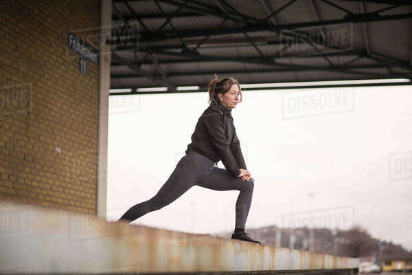 Female runner leaning forward stretching on warehouse platform ...