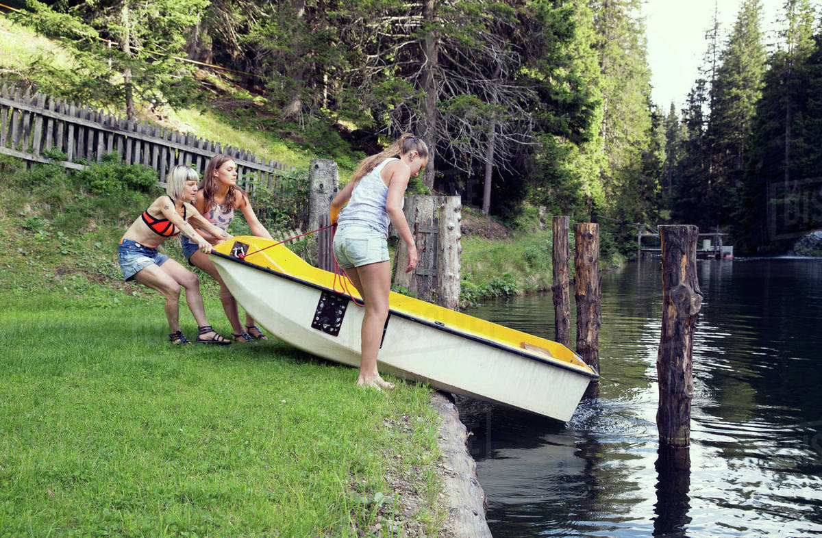 Three adult female friends launching rowing boat into lake ...
