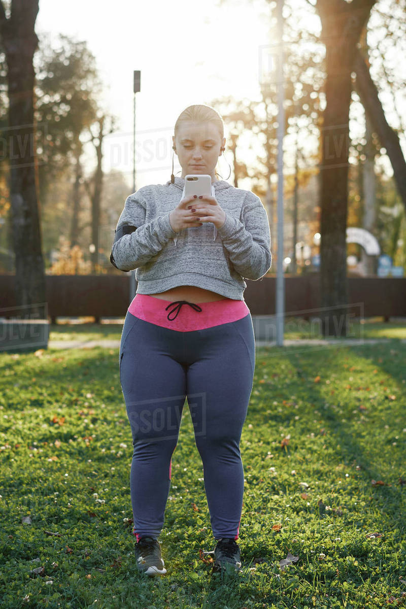 Curvaceous young woman training in park, looking at smartphone ...
