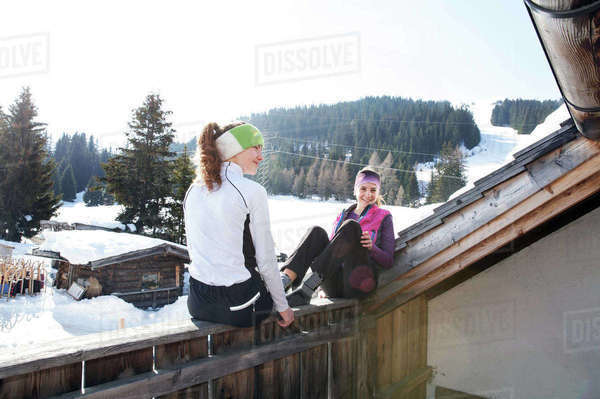 Two teenage girl skiers sitting on cabin roof terrace in snow covered ...