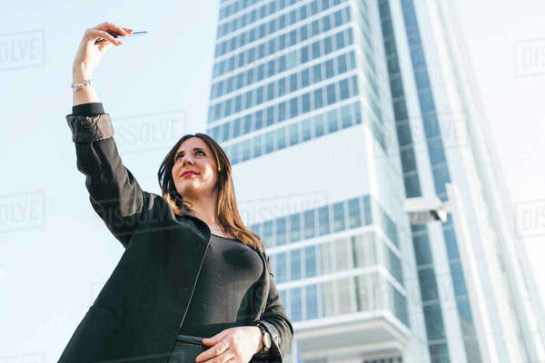 Young woman taking smartphone selfie in front of skyscraper, low angle ...