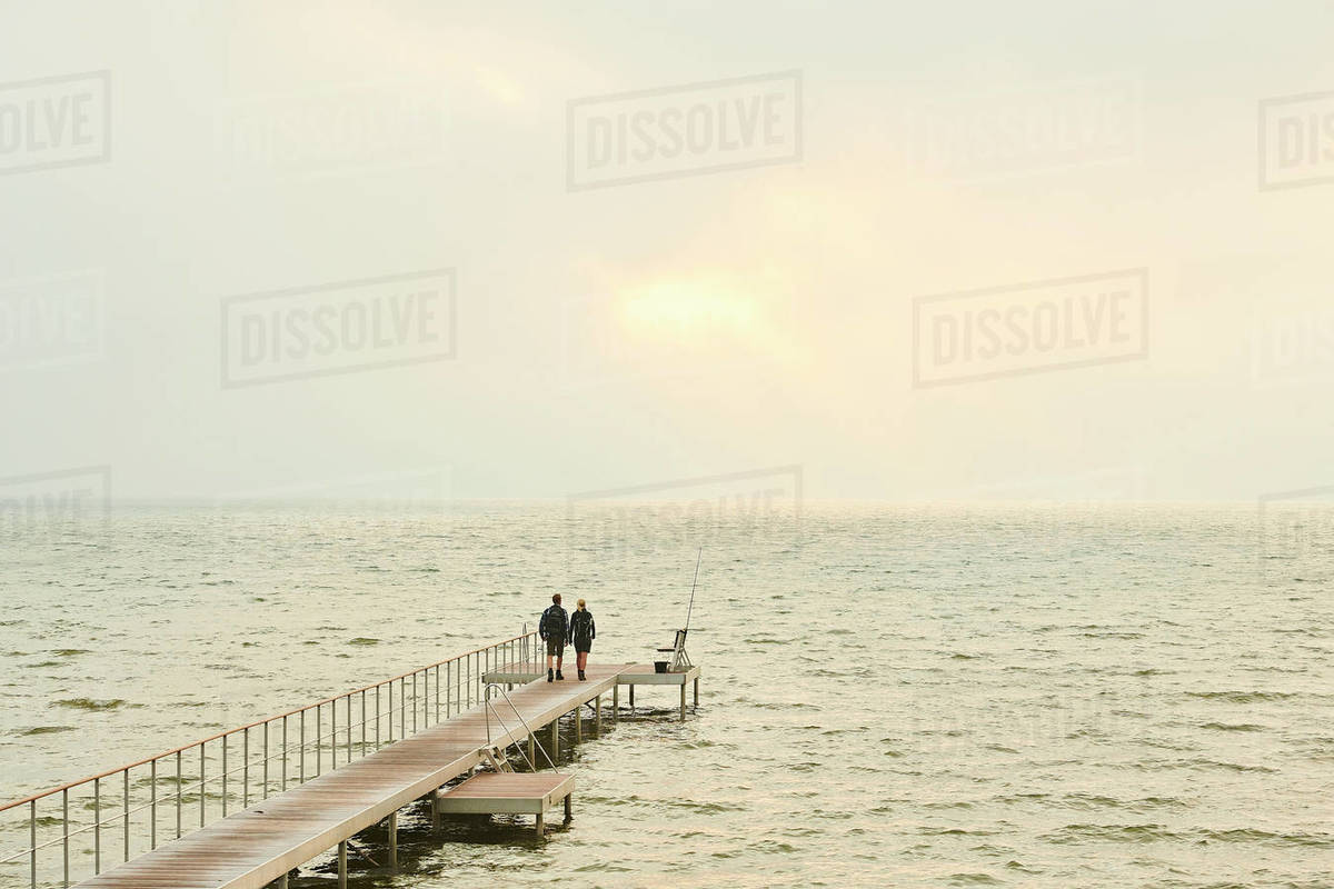 Two people standing on a jetty looking out over water to a pale sunset ...