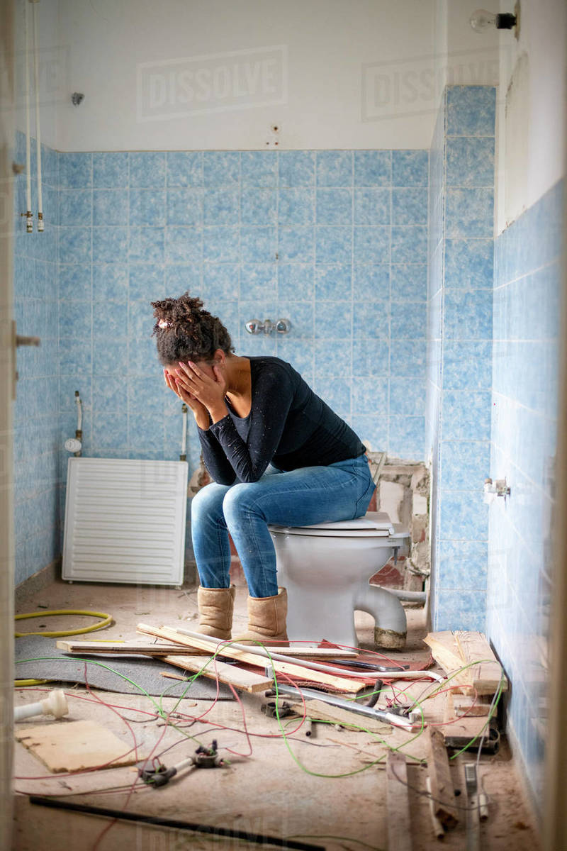 A person covers their face with their hands, seated on a toilet in a ...