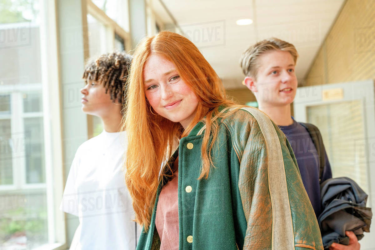 Three teenagers stand in a school corridor, smiling and looking relaxed ...