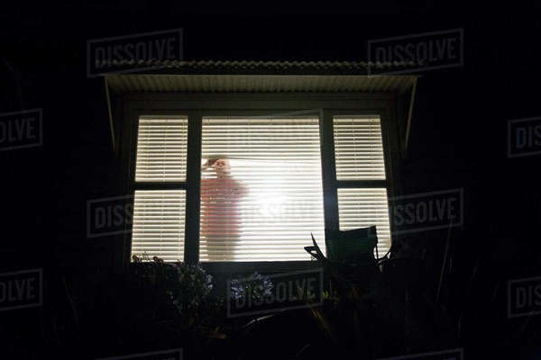 Silhouette of a person standing behind a window with blinds at night ...