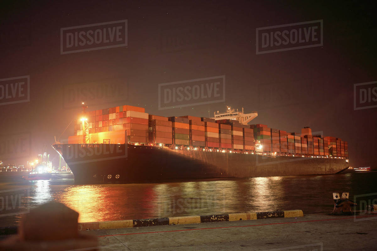 A container ship is docked at a port at night, illuminated under the ...