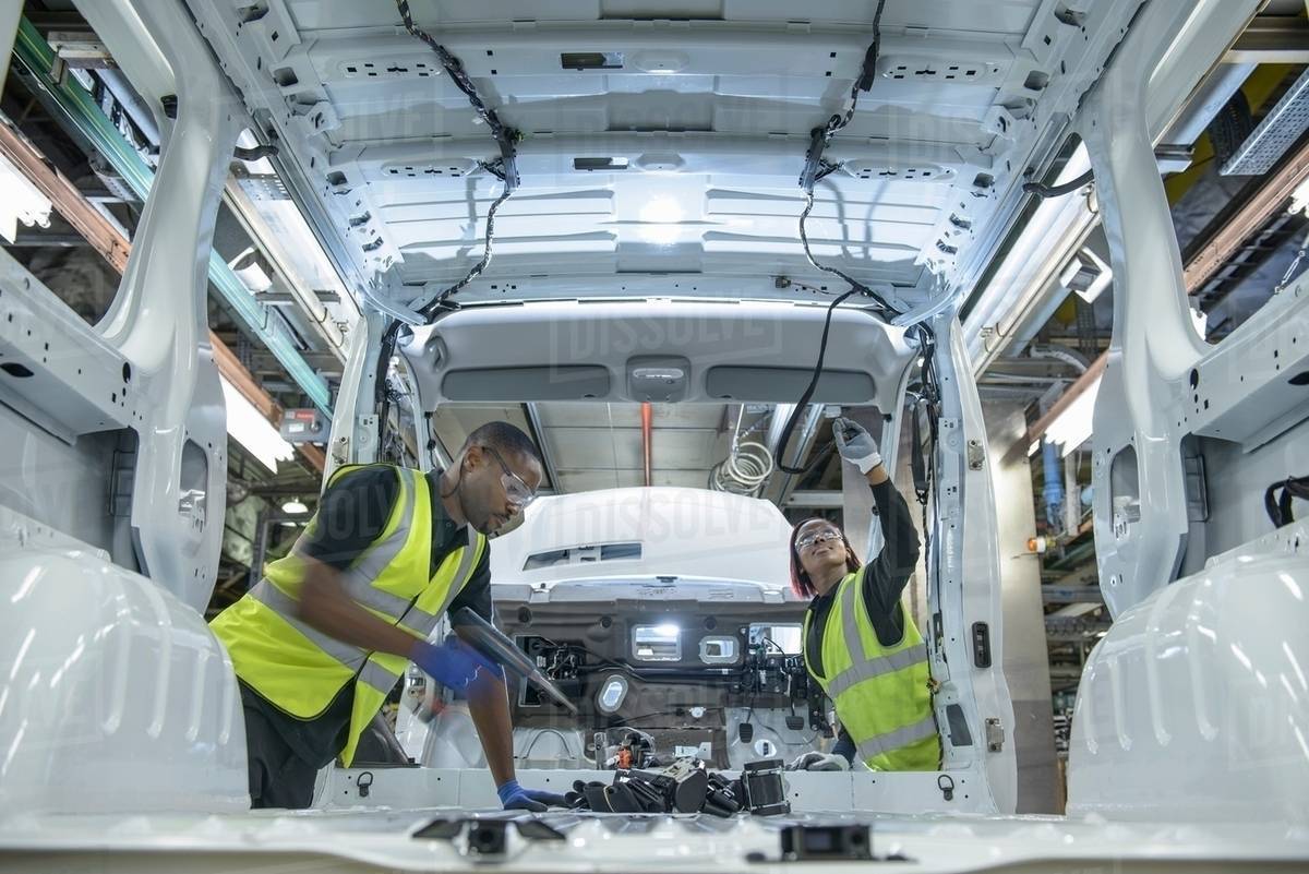 Car workers wearing high visibility jackets fitting parts on vehicle on ...