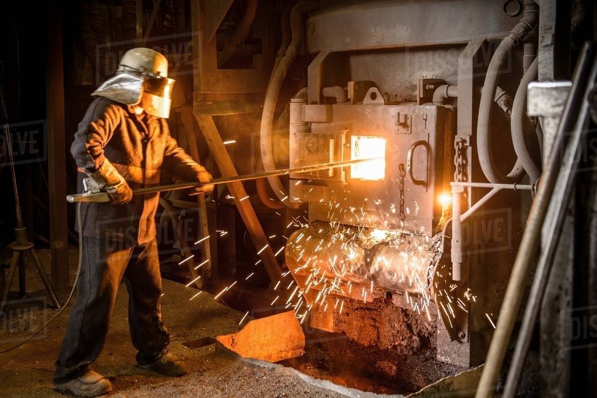Steel worker in protective clothing raking furnace in an industrial ...