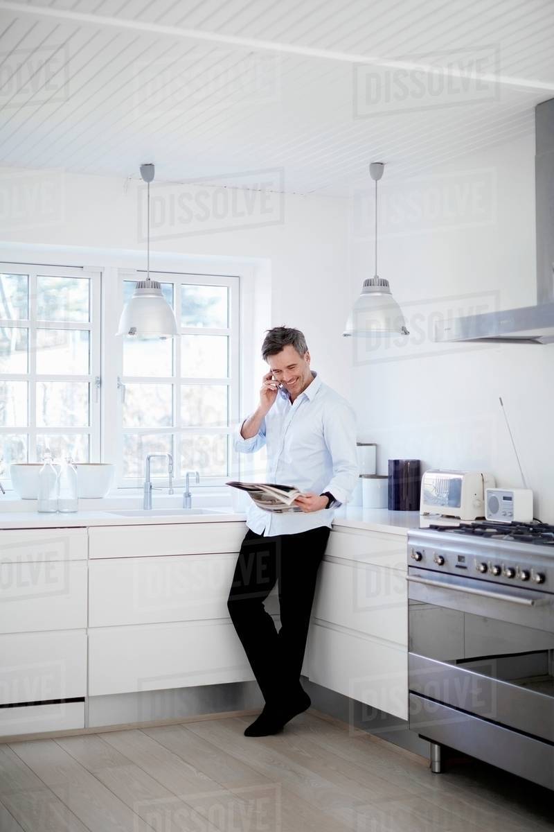 Man standing in kitchen making phone-call and reading newspaper ...
