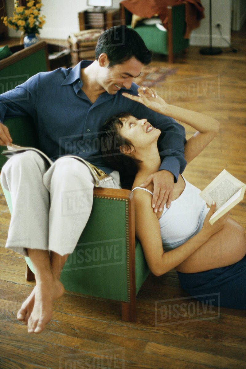 Pregnant woman sitting on floor with book, leaning back against husband ...