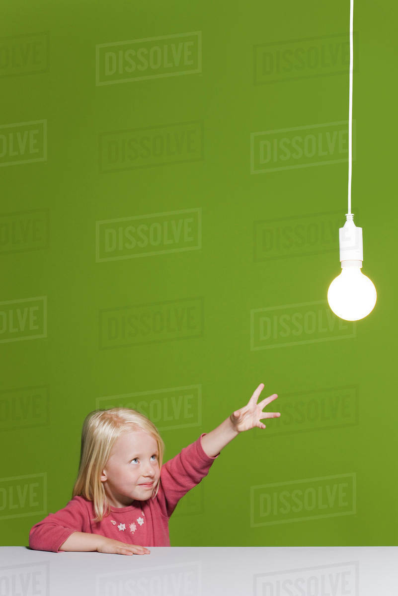 Little girl reaching for illuminated light bulb suspended overhead ...