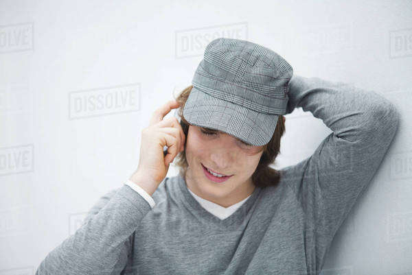 Young man in cap using cell phone, looking down, one hand behind head ...