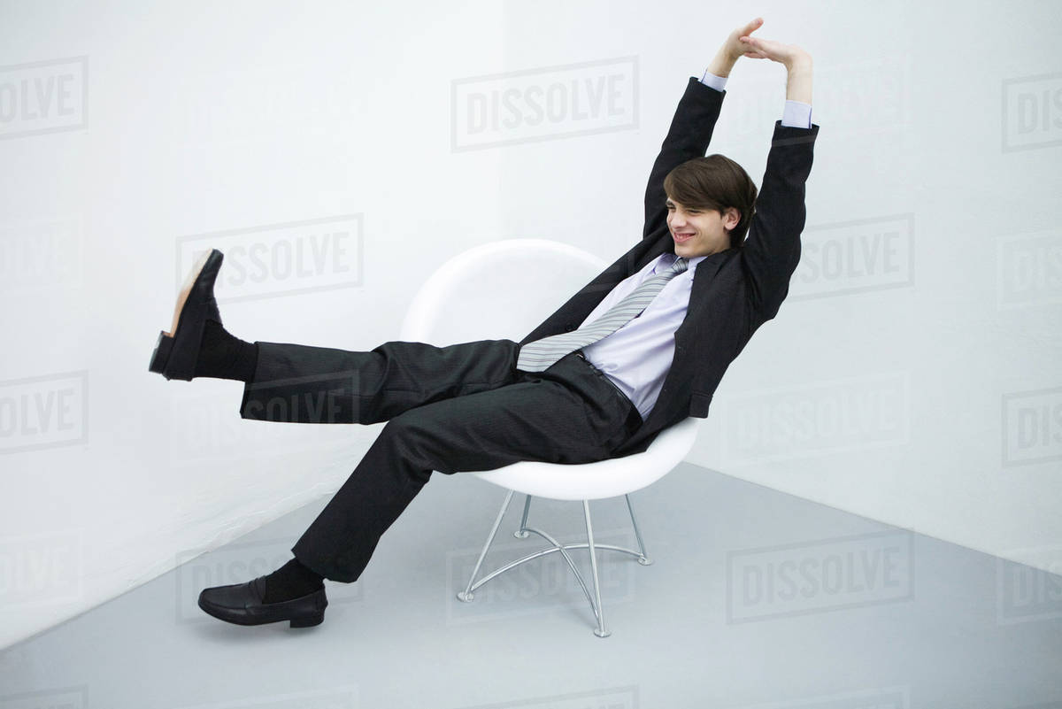 Young man in suit sitting in chair, legs outstretched, arms raised ...