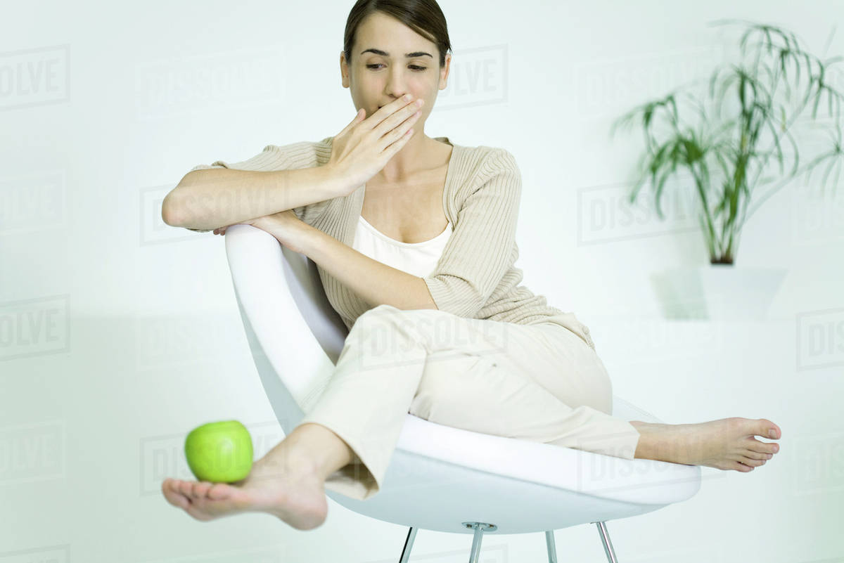 Young woman slouching in chair, balancing apple on foot, covering mouth ...