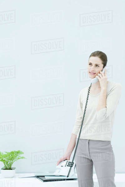 Woman standing and talking on landline phone, looking away and smiling ...