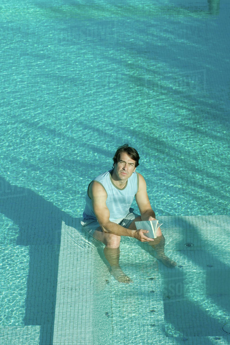 Man sitting in pool, reading, looking up at camera - Stock Photo - Dissolve