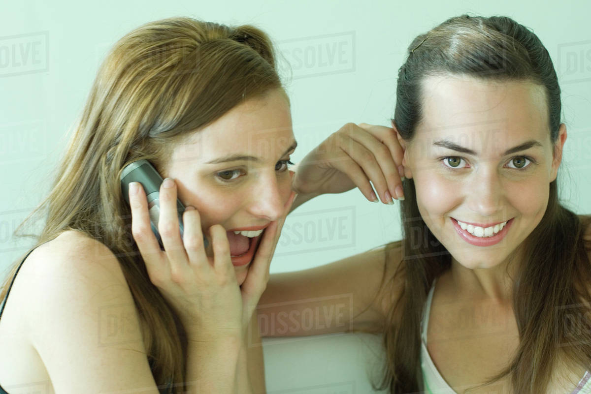 Two young friends smiling together, using cell phone, one looking at ...