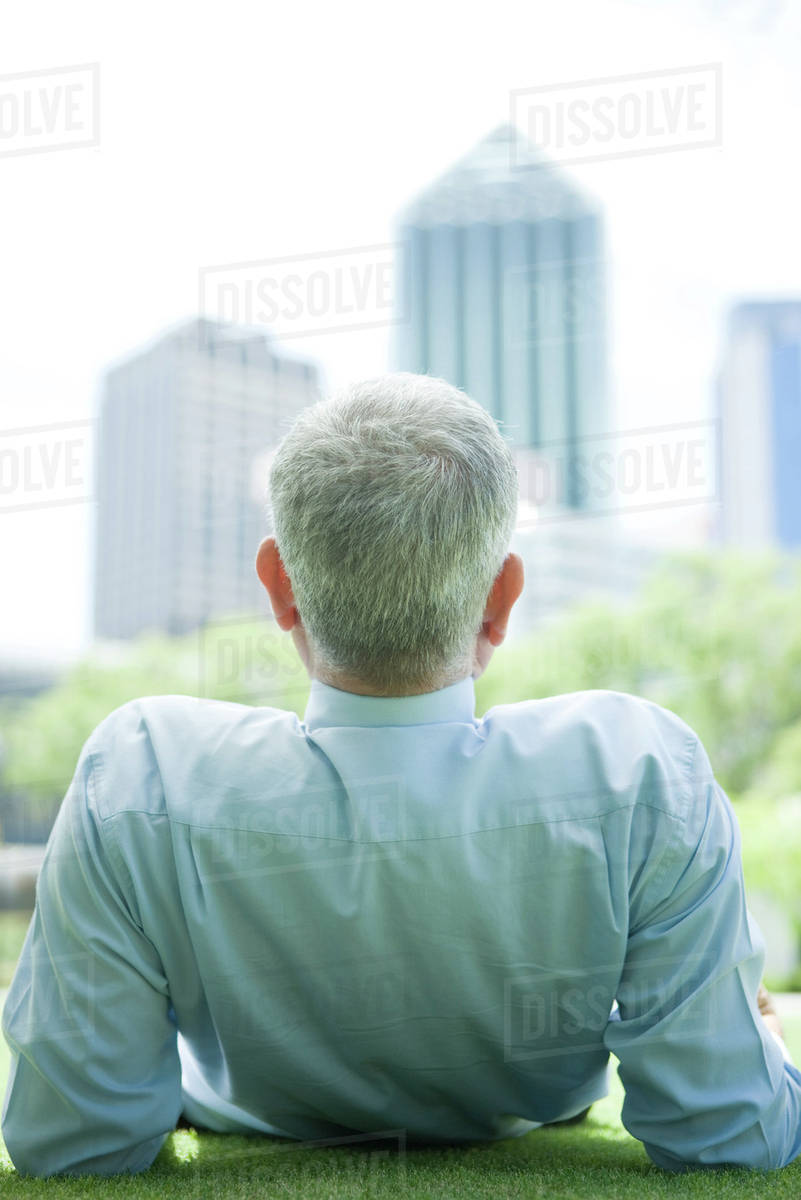 Mature man reclining on ground, looking at urban scene, rear view ...