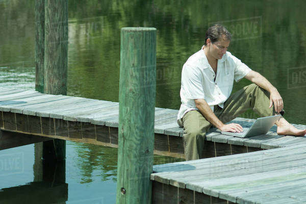 Man sitting on dock, using laptop computer, looking down, full length ...